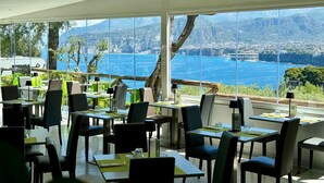 Restaurante com mesas e cadeiras dispostas perto de amplas janelas com vista para a paisagem costeira.