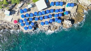Vista aérea de uma área costeira com vários guarda-sóis azuis dispostos em fileiras, um edifício branco e terreno rochoso próximo à água.