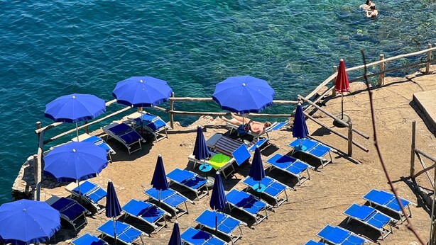 Uma praia com guarda-sóis azuis, espreguiçadeiras e uma passarela de madeira.