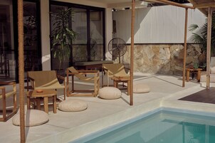 Área de piscina externa com espreguiçadeiras, uma mesa e um muro de pedra.