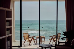 Quarto com uma janela grande de frente para o mar, duas cadeiras de madeira, uma mesinha e uma estante de livros.