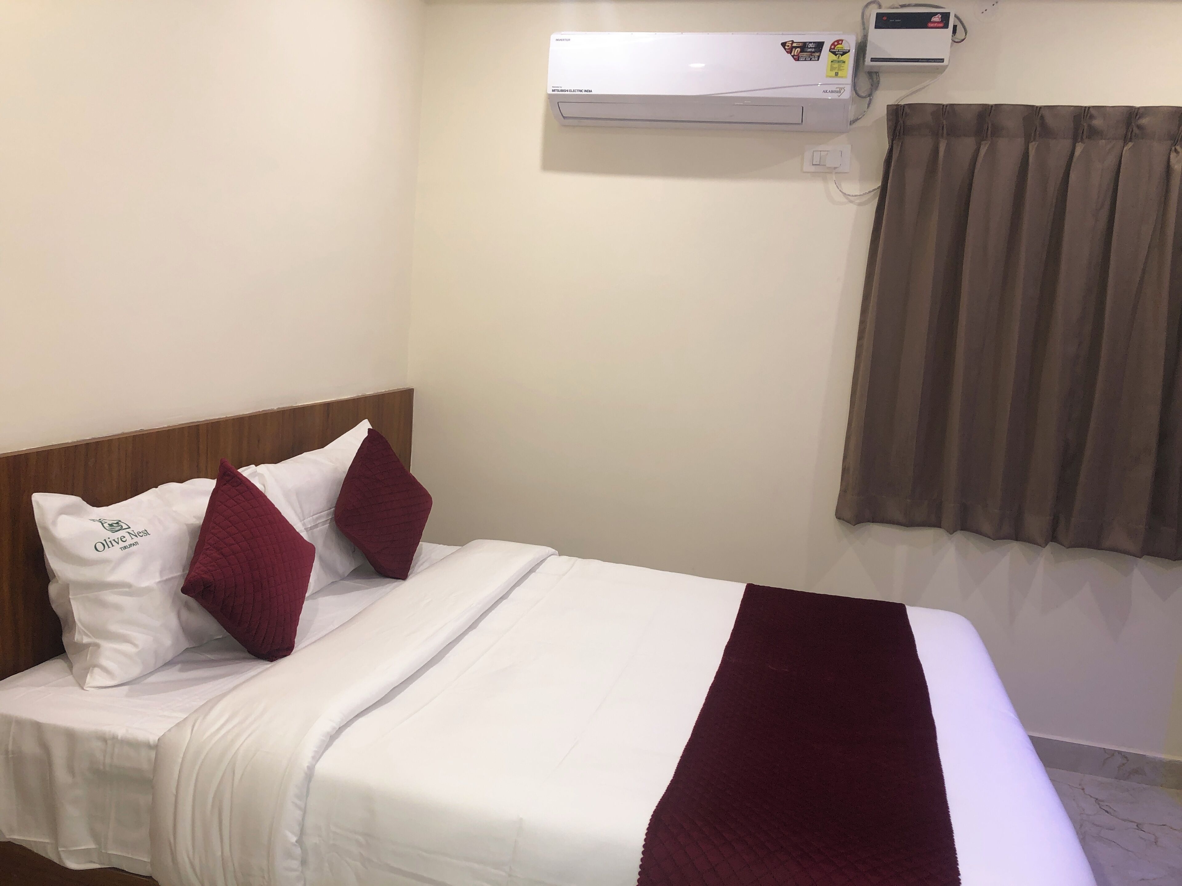Quarto de hotel com cama, roupa de cama branca, cabeceira marrom, ar condicionado fixado na parede, janela com cortinas e uma pequena mesa com abajur.
