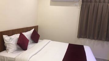 Quarto de hotel com cama, roupa de cama branca, cabeceira marrom, ar condicionado fixado na parede, janela com cortinas e uma pequena mesa com abajur.
