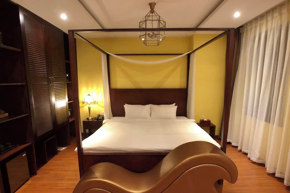Quarto de hotel com cama de dossel, piso de madeira, mesa de cabeceira com luminária e guarda-roupa.