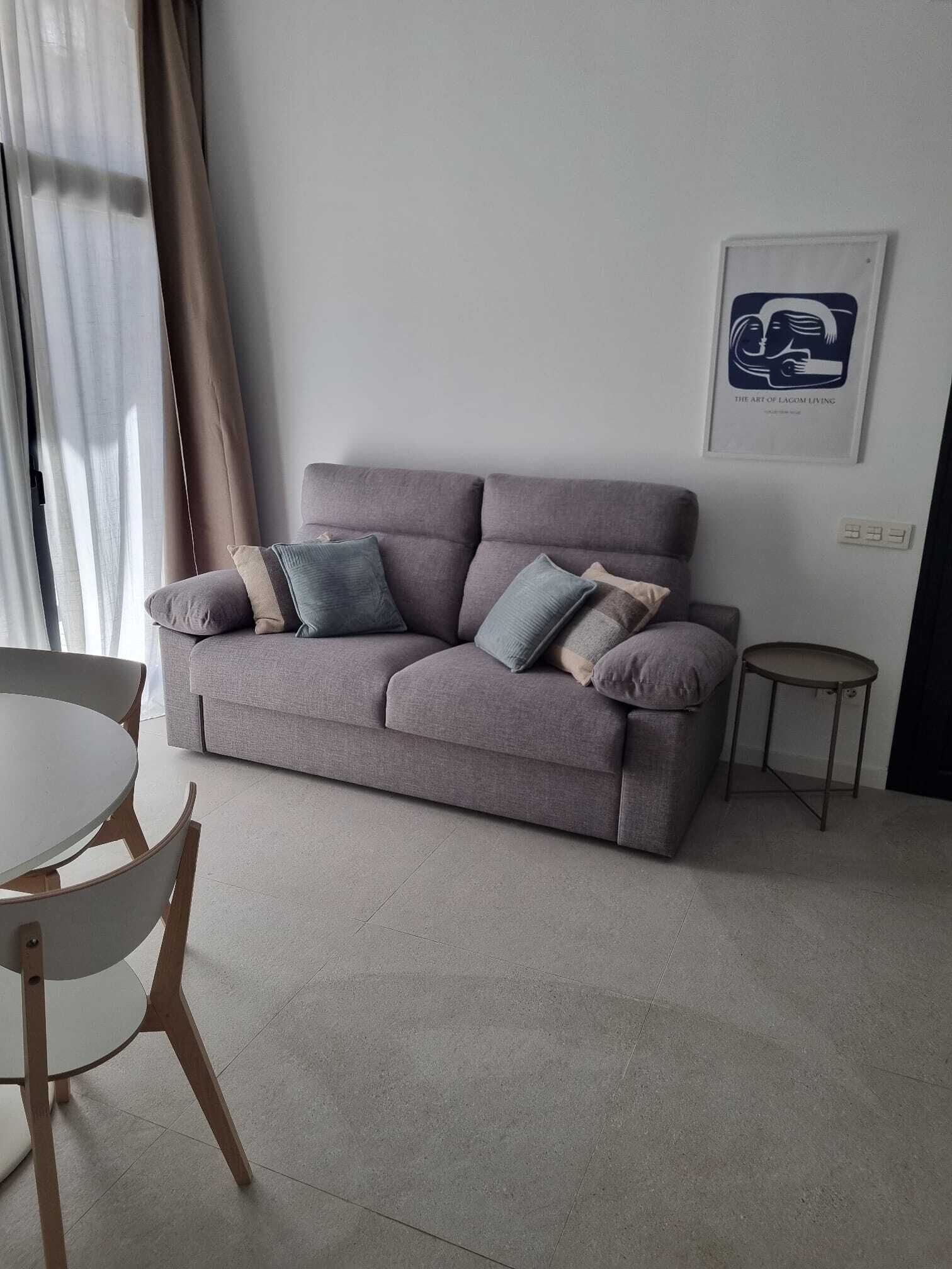 Uma sala de estar moderna com um sofá cinzento, uma mesa de jantar redonda e uma pequena mesa lateral.
