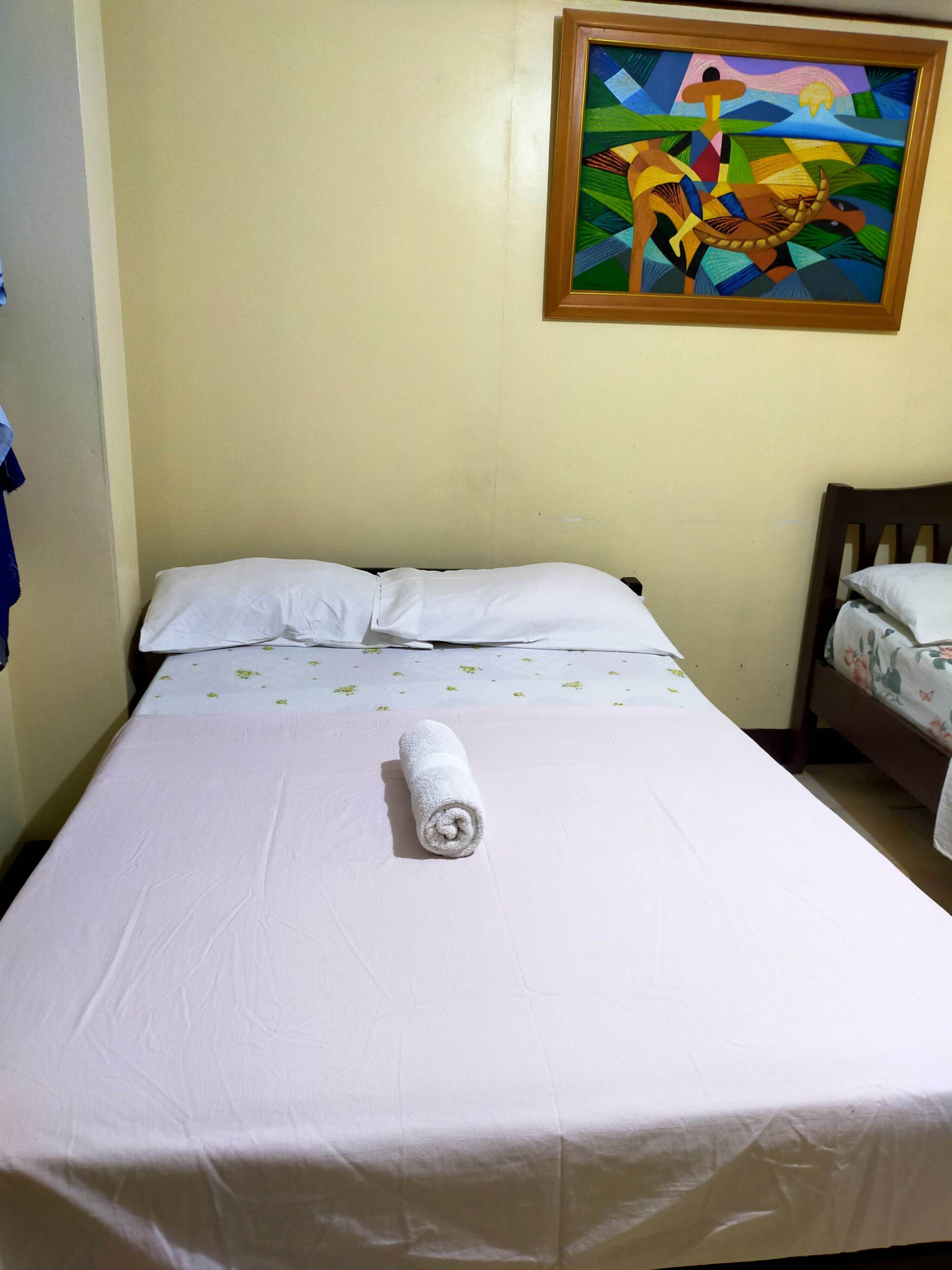 Uma cama de solteiro com uma toalha enrolada, um quadro na parede e parte de outra cama à direita.
