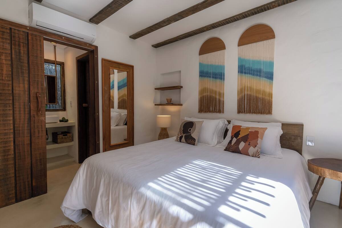 Quarto com cama grande, vigas de madeira no teto, guarda-roupa de madeira, espelho e prateleira na parede com uma tigela.