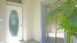 Quarto de hotel com porta de vidro fosco, uma pequena janela e um vaso de planta perto do elevador.