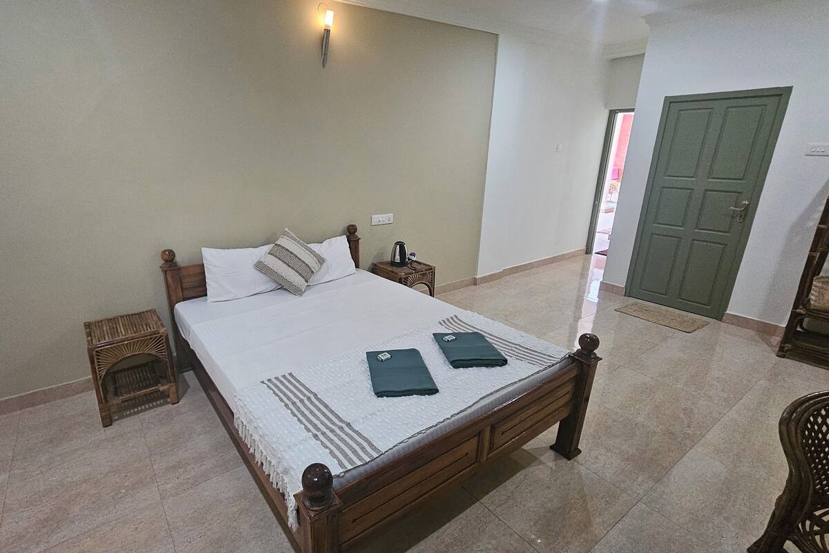Uma cama bem arrumada com almofadas e um cobertor, uma mesa de cabeceira com chaleira, e uma porta verde em um quarto com piso de azulejos.