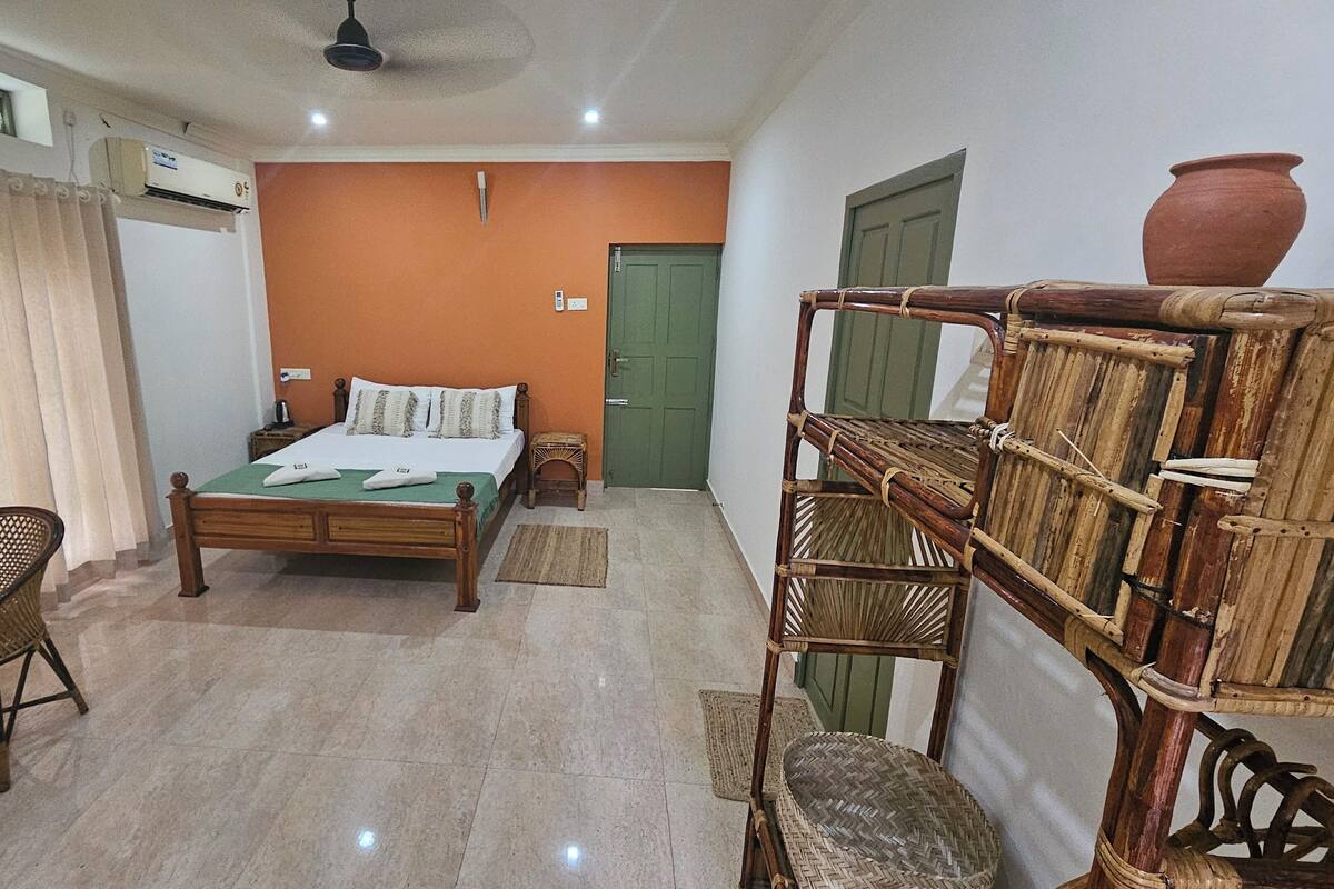 Quarto com uma cama, uma cadeira, uma mesinha, uma cadeira de fibra natural e uma porta verde.