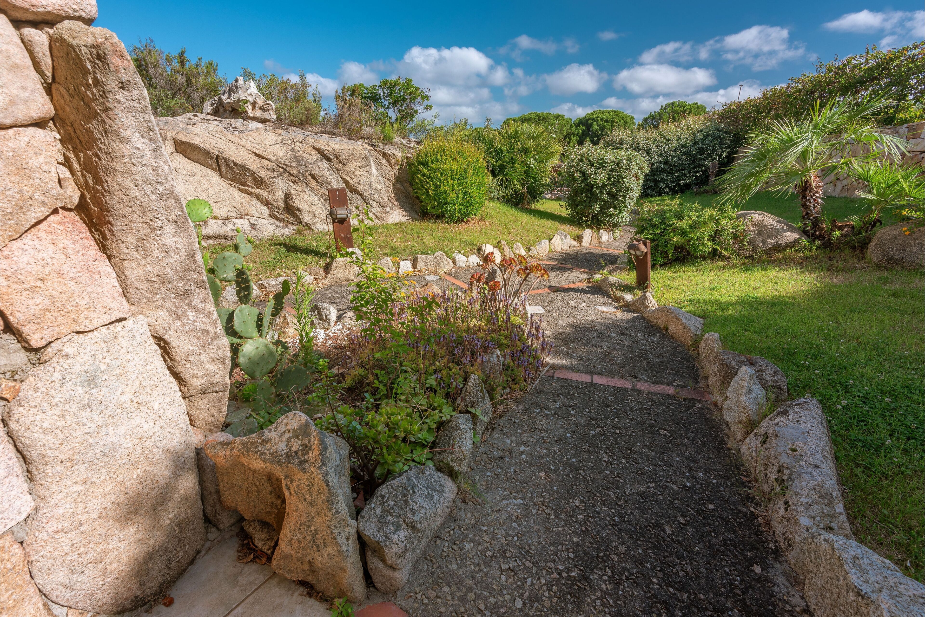Um caminho de jardim ladeado por pedras grandes e diversas plantas, que leva a um muro de pedra.