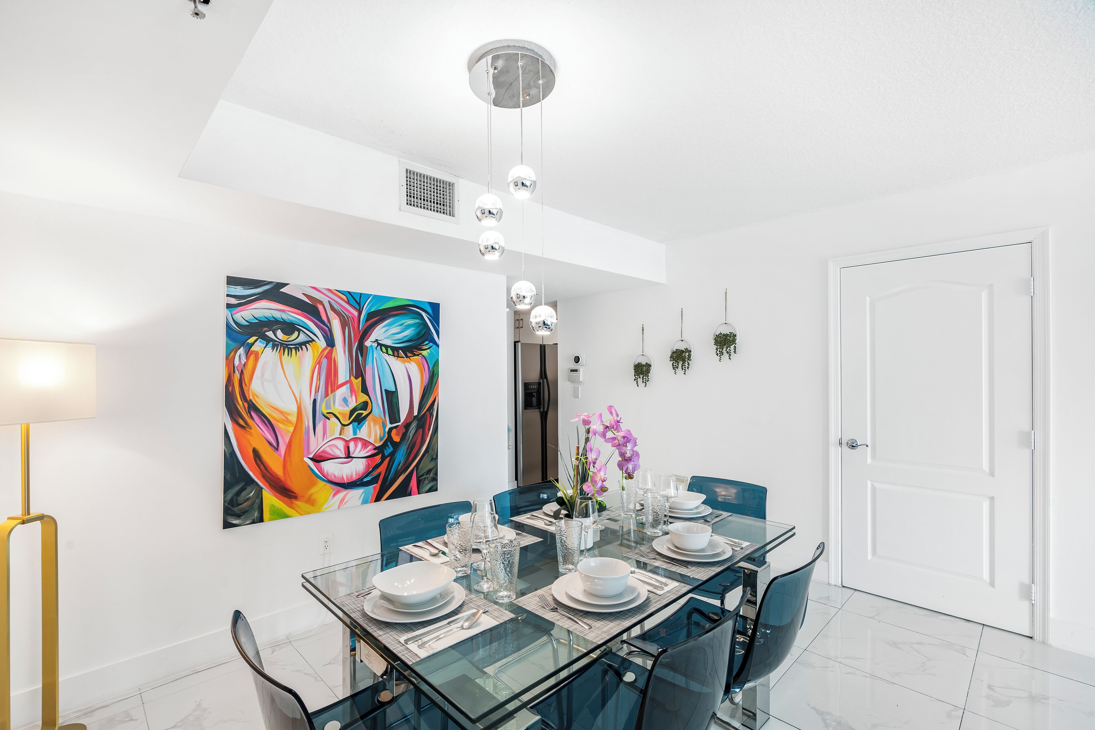 Uma sala de jantar moderna com mesa de vidro, cadeiras azuis, paredes brancas e uma grande pintura abstrata.