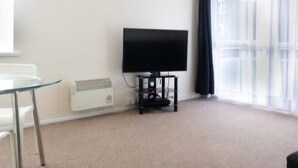 Uma sala de estar com televisão de tela plana em um suporte, radiador branco, mesa de vidro e uma janela grande com cortinas.
