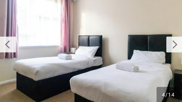 Quarto de hotel com duas camas de solteiro, uma janela com cortinas, uma mesa de cabeceira e uma luminária.