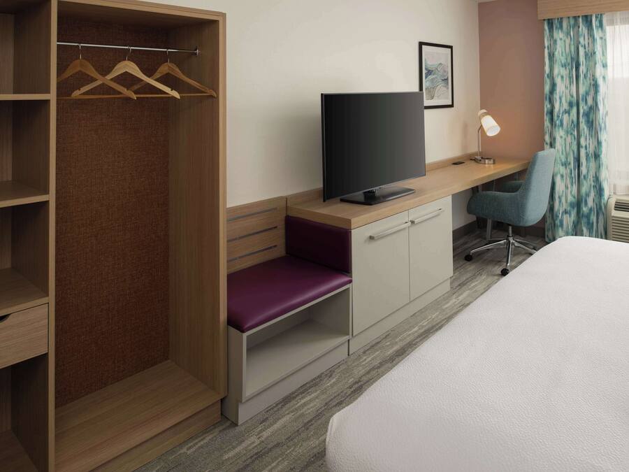 Quarto de hotel com cama, secretária com televisão, uma cadeira, guarda-roupa e janela com cortinas.