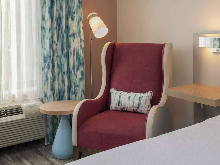 Quarto de hotel com poltrona vermelha, mesa redonda pequena, mesa de cabeceira, chuveiro embutido na parede e janela com cortinas translúcidas.