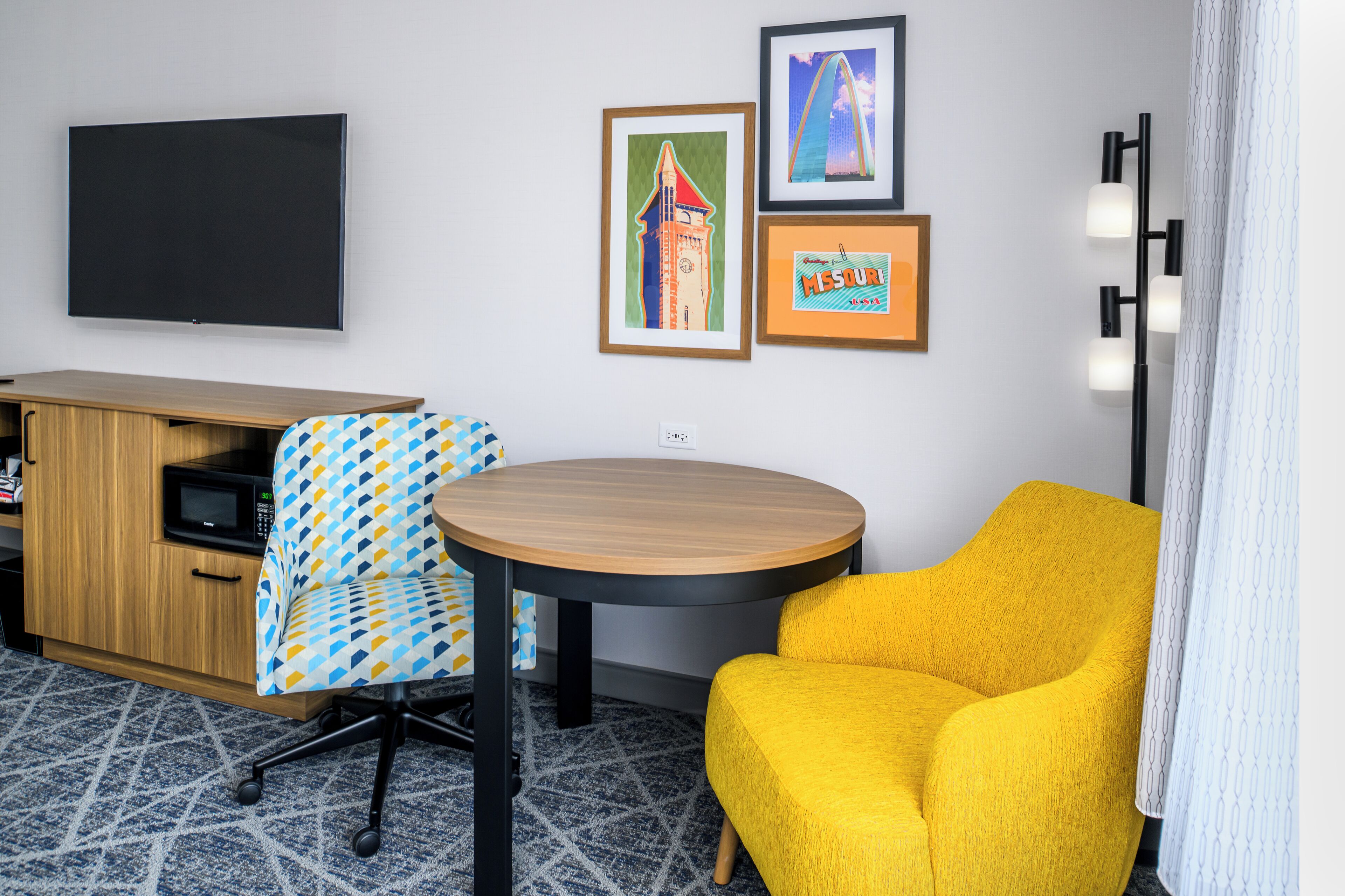 Quarto com escrivaninha de madeira, poltrona amarela, mesa redonda e televisão fixada na parede.