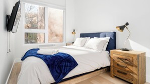 Quarto com cama, mesa de cabeceira com abajur, TV fixada na parede e janela com vista para árvores.