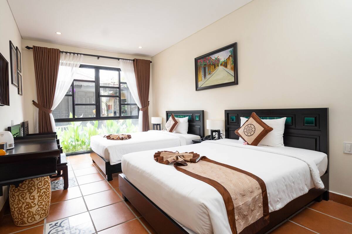 Quarto de hotel com duas camas, uma escrivaninha, uma cadeira e uma janela com cortinas.