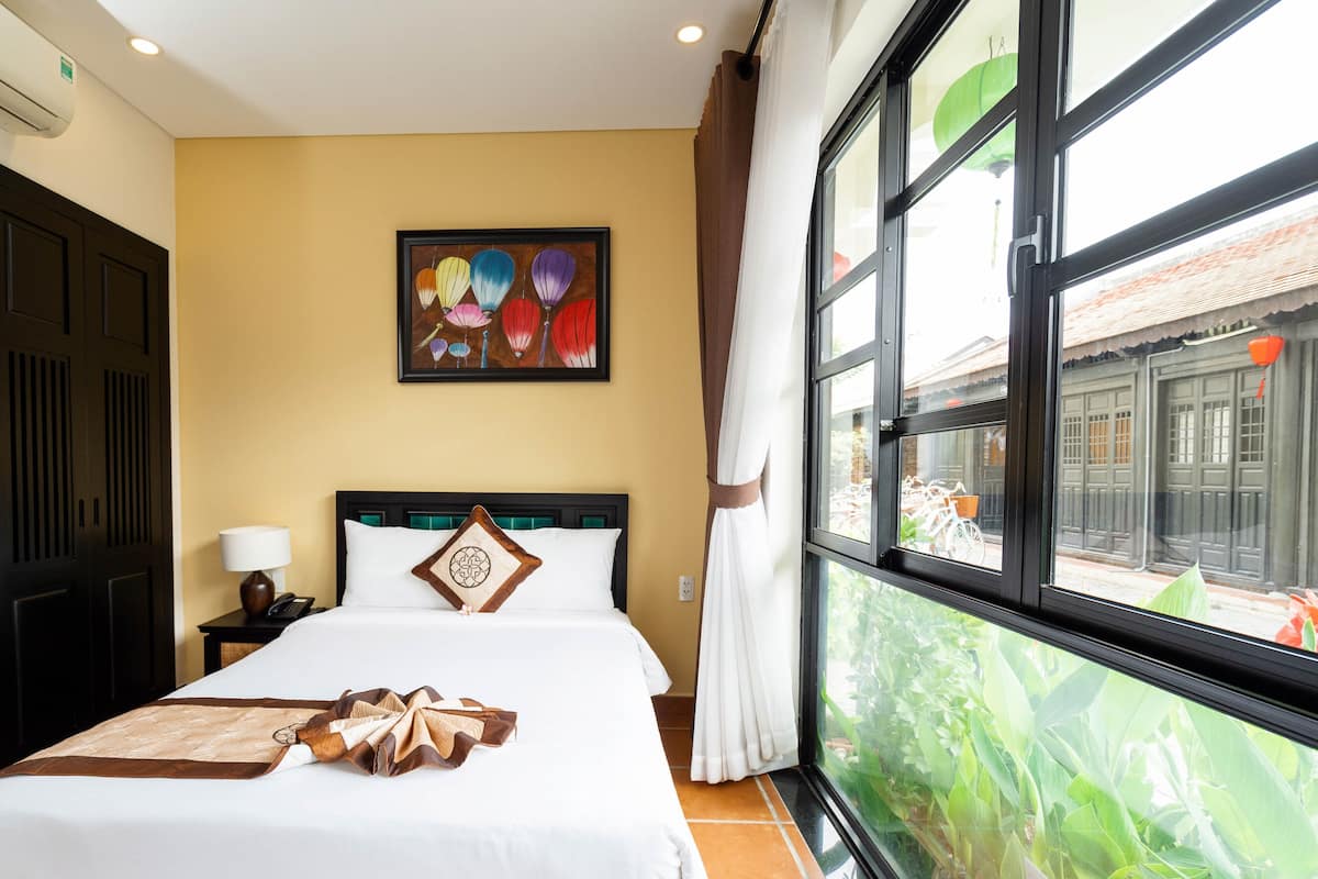 Quarto de hotel com cama, mesa de cabeceira, abajur, pintura e janela com vista para áreas verdes e edifícios.