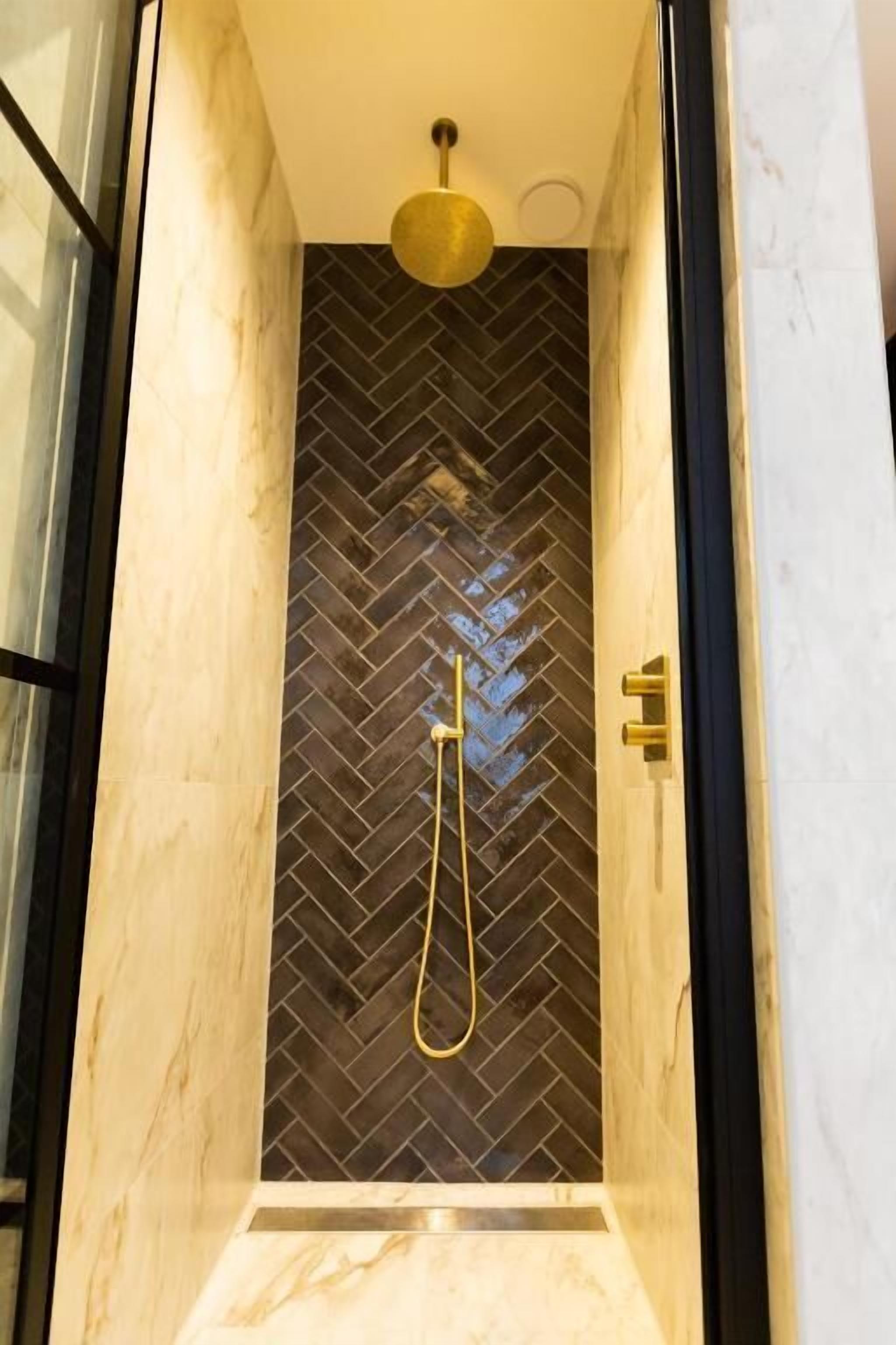 Chuveiro com parede em padrão herringbone, um chuveiro dourado e uma porta de madeira com maçanetas de latão.