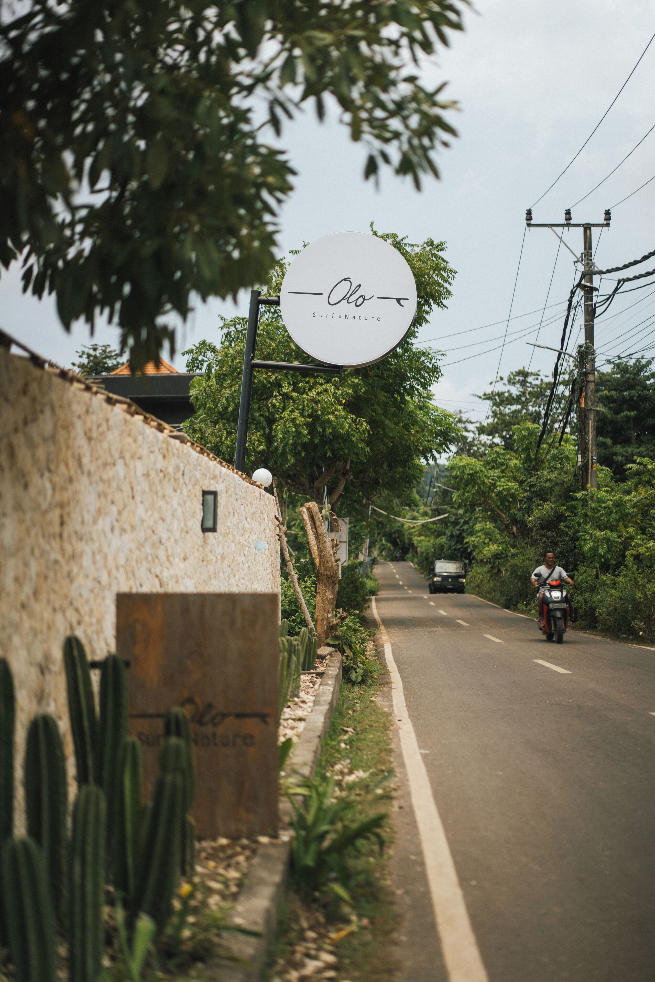 Uma estrada com uma placa indicando 'Olo SurfNature' e um motociclista ao longe.
