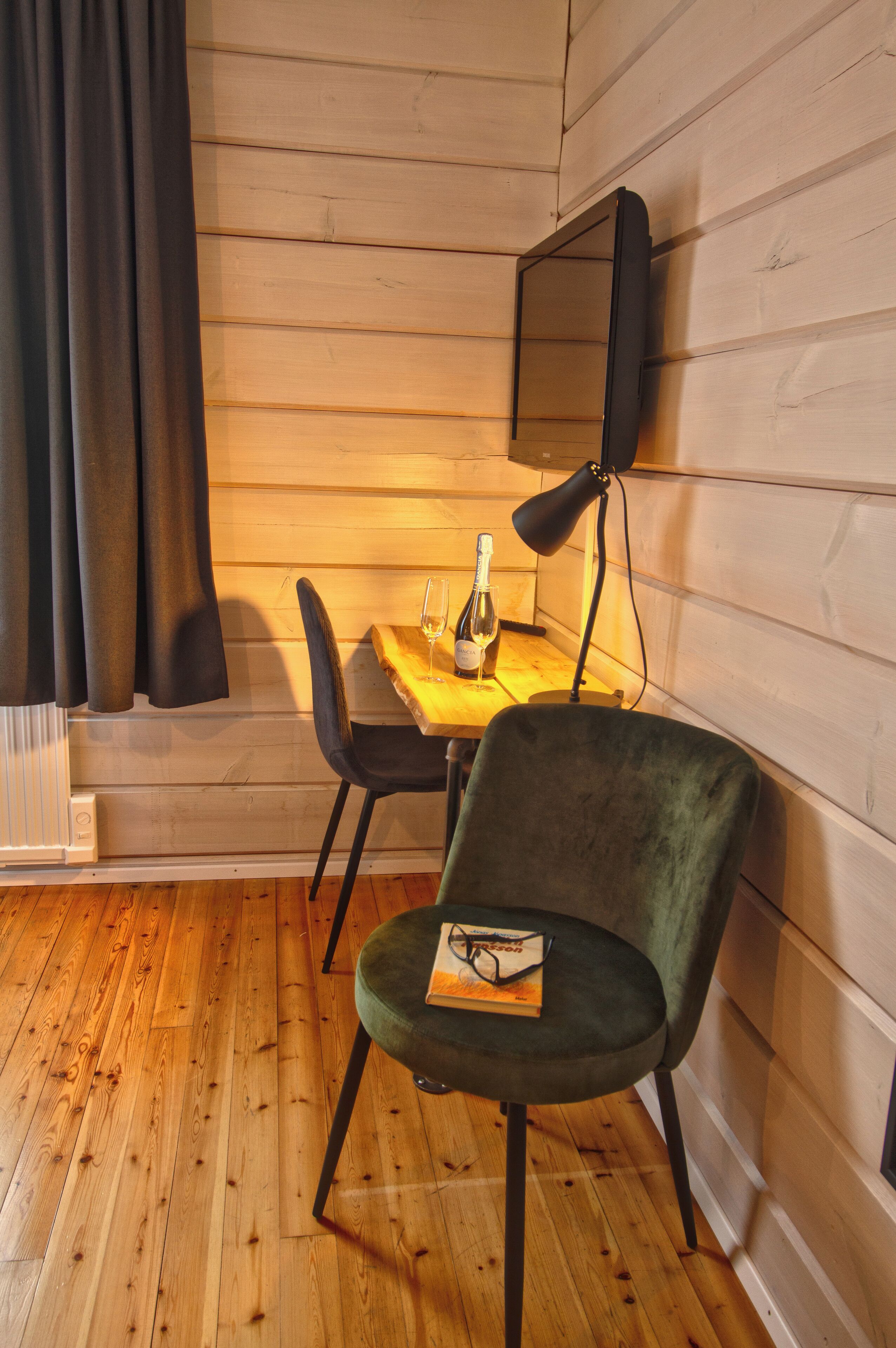 Quarto aconchegante com paredes e piso de madeira, uma mesa com uma garrafa e copos, uma cadeira e um abajur.