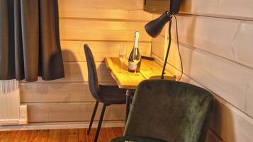 Quarto aconchegante com paredes e piso de madeira, uma mesa com uma garrafa e copos, uma cadeira e um abajur.