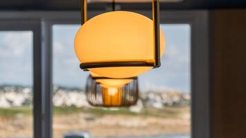 Um close-up de uma luminária pendente com luz quente, uma janela com vista para um estacionamento e utensílios de cozinha em uma bancada.