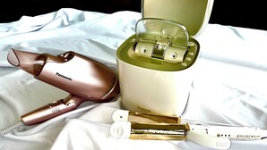 Um secador de cabelo Panasonic, uma escova de cabelo branca e dourada, e um alisador de cabelo branco e dourado sobre uma superfície de tecido branca.