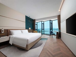Quarto de hotel moderno com uma cama grande, uma escrivaninha, uma cadeira e uma ampla janela com vista para um lago e montanhas.