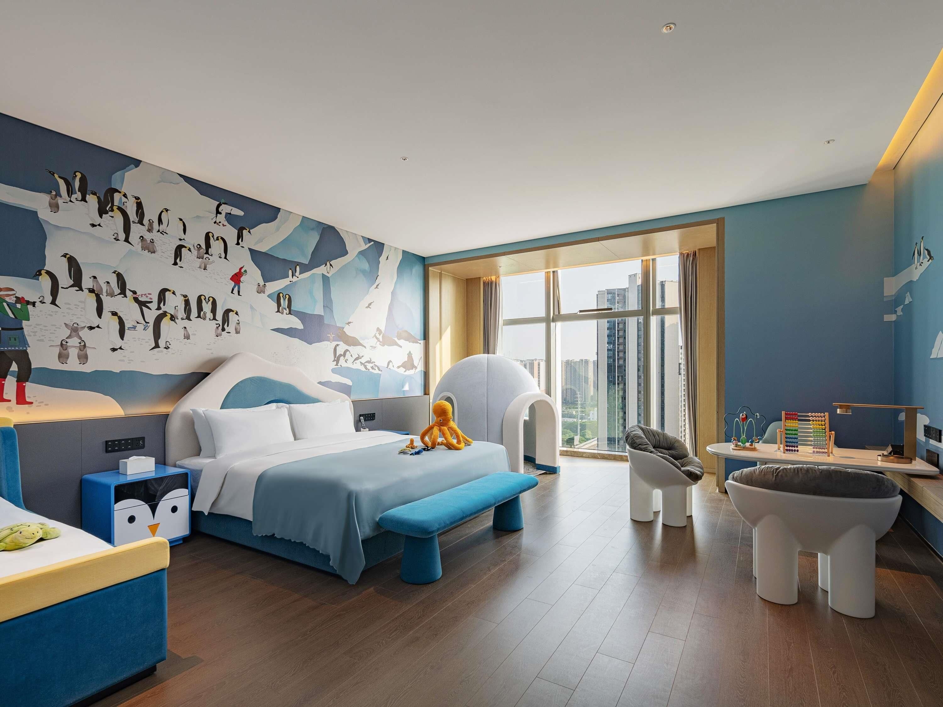 Quarto de hotel moderno com uma grande janela, cama com cabeceira azul, escrivaninha com cadeira e um mural na parede com pinguins.