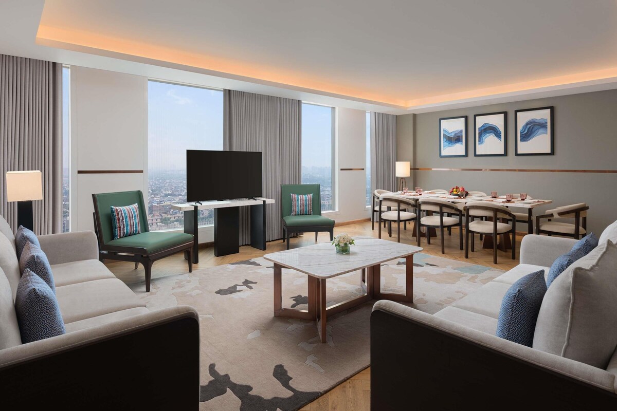 Quarto de hotel moderno com televisão de tela plana, poltronas verdes, mesa de jantar e vista para a cidade.