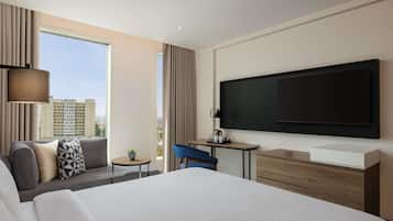 Quarto de hotel moderno com uma cama grande, um sofá, uma mesa pequena, uma televisão e uma janela com vista para a cidade.