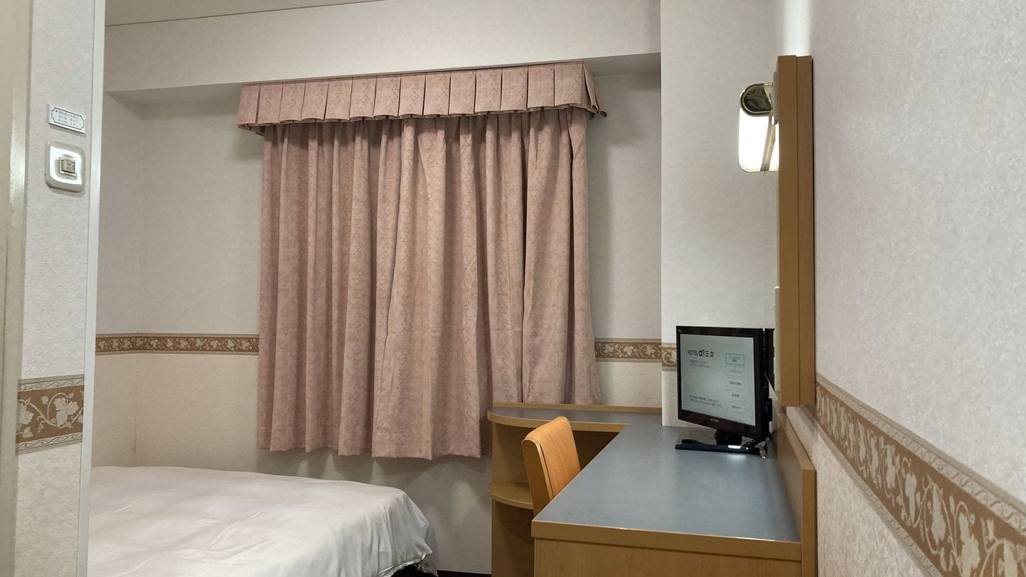 Quarto de hotel com cama, escrivaninha e computador, além de janela com cortinas.