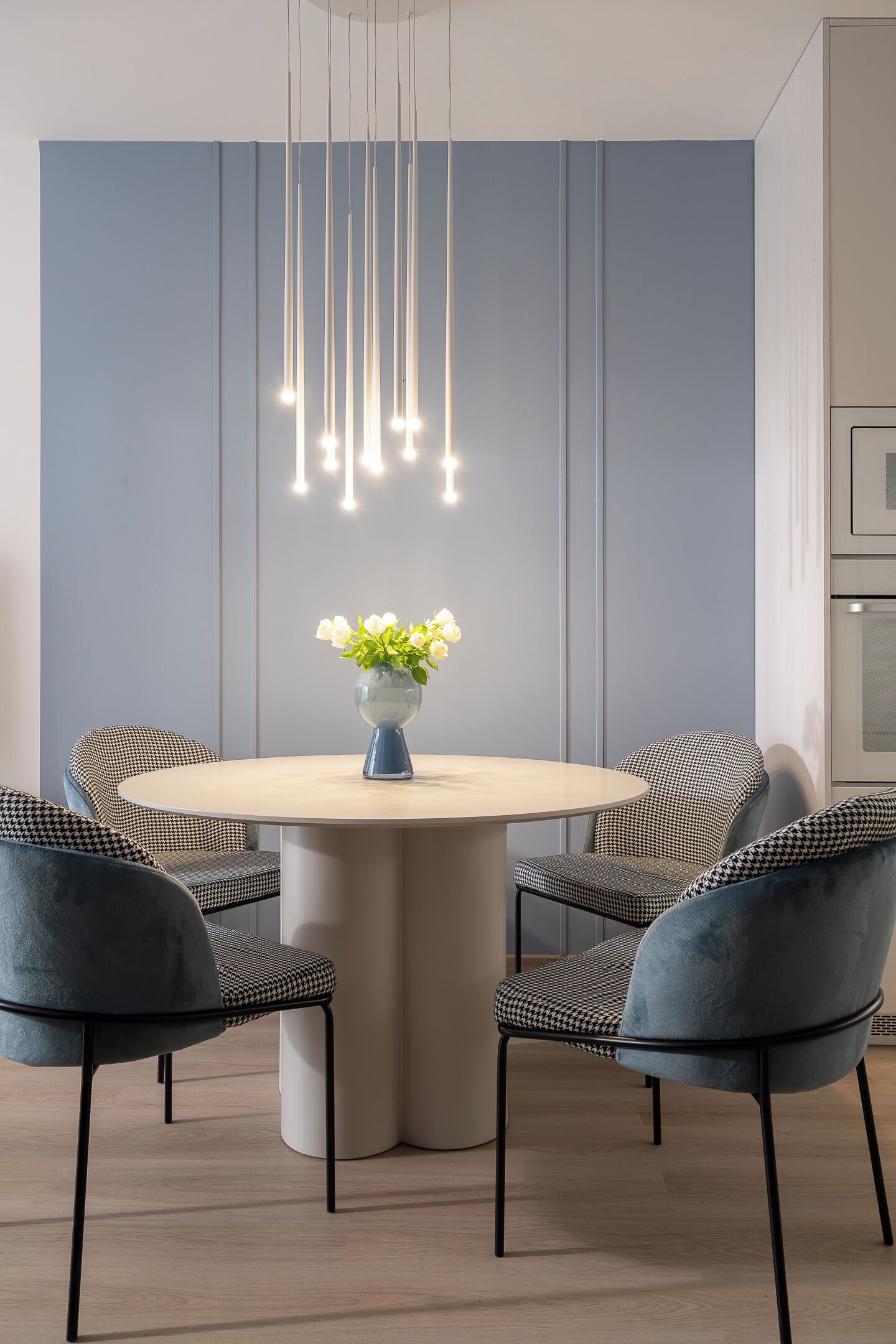 Uma área de refeições com uma mesa redonda, quatro cadeiras, um vaso com flores e um lustre.