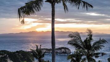 Uma palmeira em primeiro plano, com vista para o oceano e montanhas ao longe durante o pôr do sol.