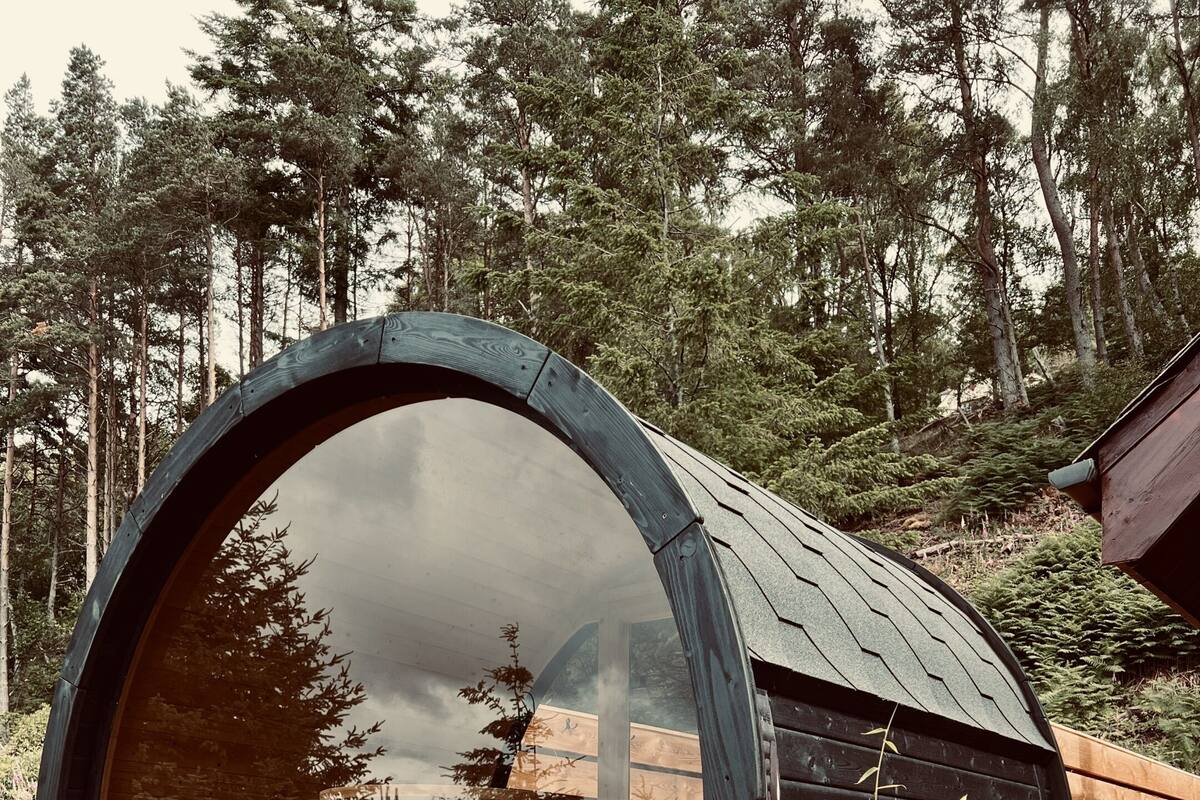 Uma estrutura cilíndrica com superfície refletora, rodeada por elementos de madeira e vegetação.