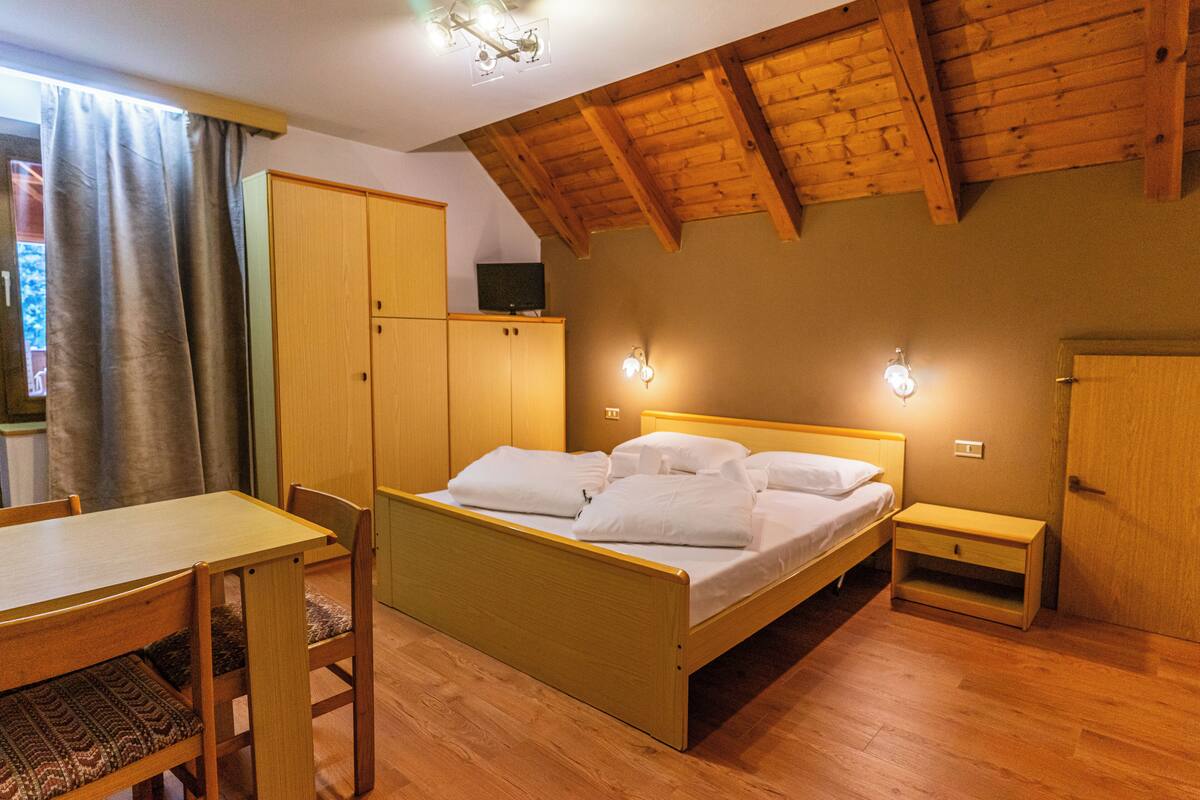 Quarto com cama de madeira, escrivaninha de madeira, uma cadeira, guarda-roupa e mesa de cabeceira pequena.