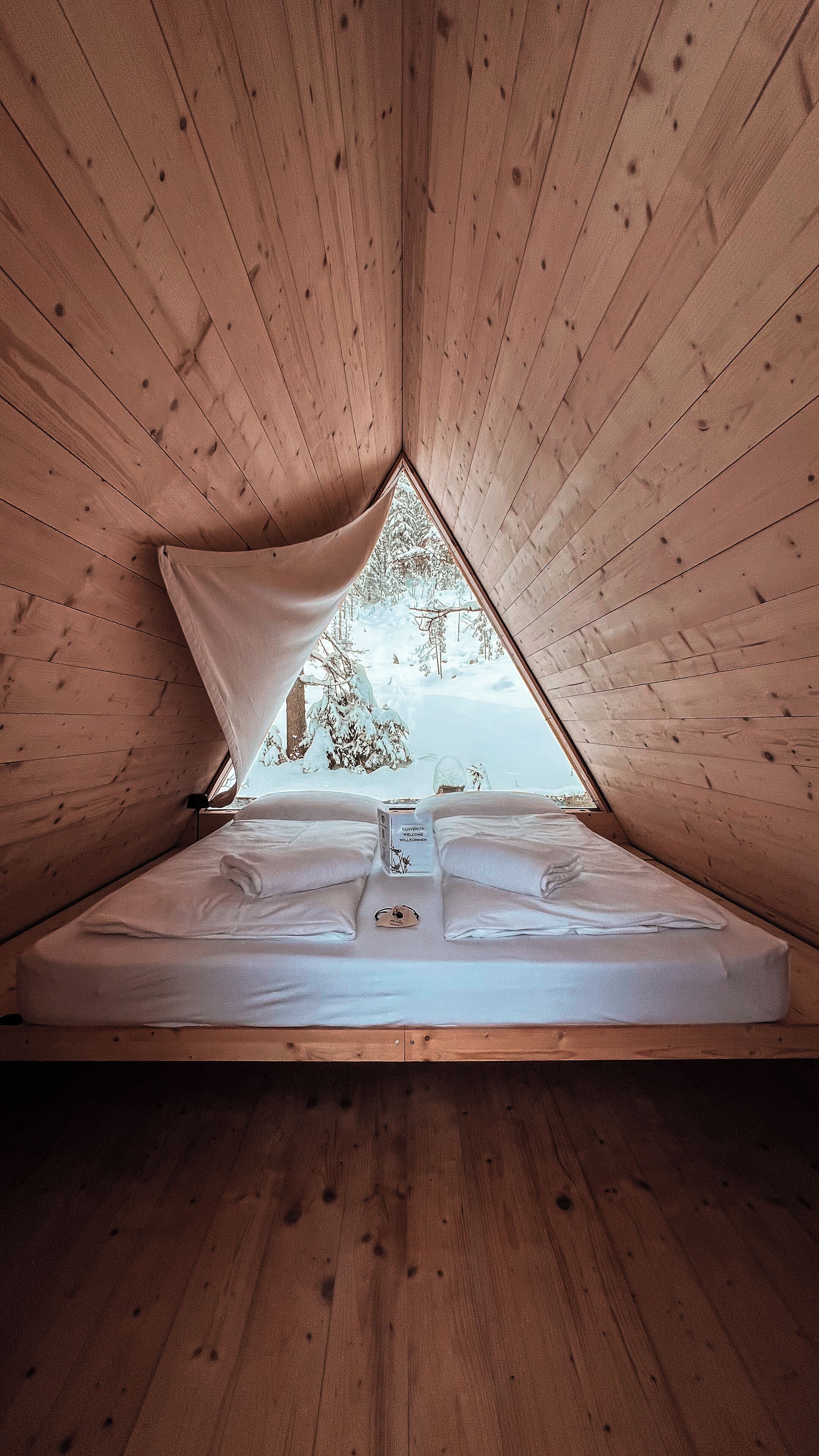 Um quarto de madeira acolhedor e triangular, com uma cama e janela com vista para uma paisagem nevada.