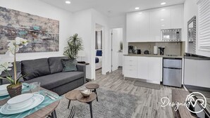 Um apartamento moderno com um sofá cinzento, uma mesa de jantar com pratos, uma cozinha com armários brancos e um grande quadro abstrato na parede.