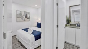Quarto de hotel moderno com uma cama bem arrumada, uma mesa de cabeceira com abajur, um quadro na parede e uma pequena planta no móvel.