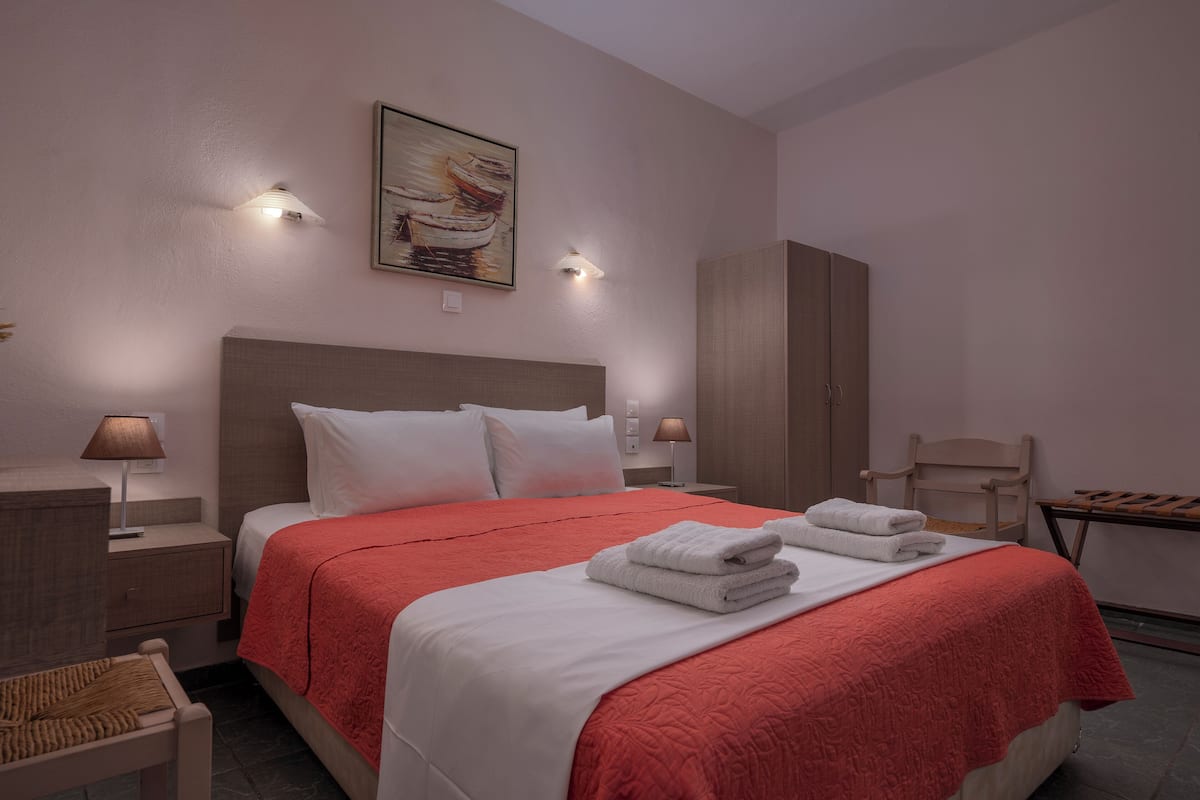 Uma cama bem arrumada com colcha vermelha e branca, duas luminárias de cabeceira, um guarda-roupa de madeira e um quadro na parede.
