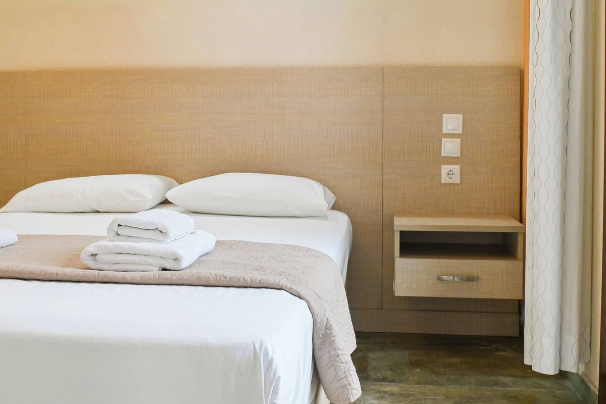 Cama bem arrumada com lençóis brancos, manta bege e toalha dobrada em uma mesa de cabeceira de madeira com gaveta.