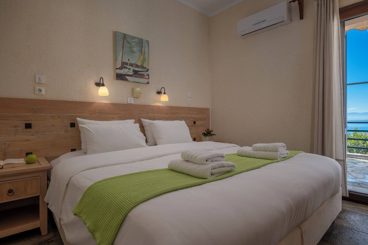 Uma cama bem arrumada com roupa de cama branca e um cobertor verde, uma cabeceira de madeira e um quadro de um veleiro na parede.