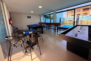 Mesa de bilhar, cadeiras e mesa de futebol de mesa numa sala bem iluminada com grandes janelas.