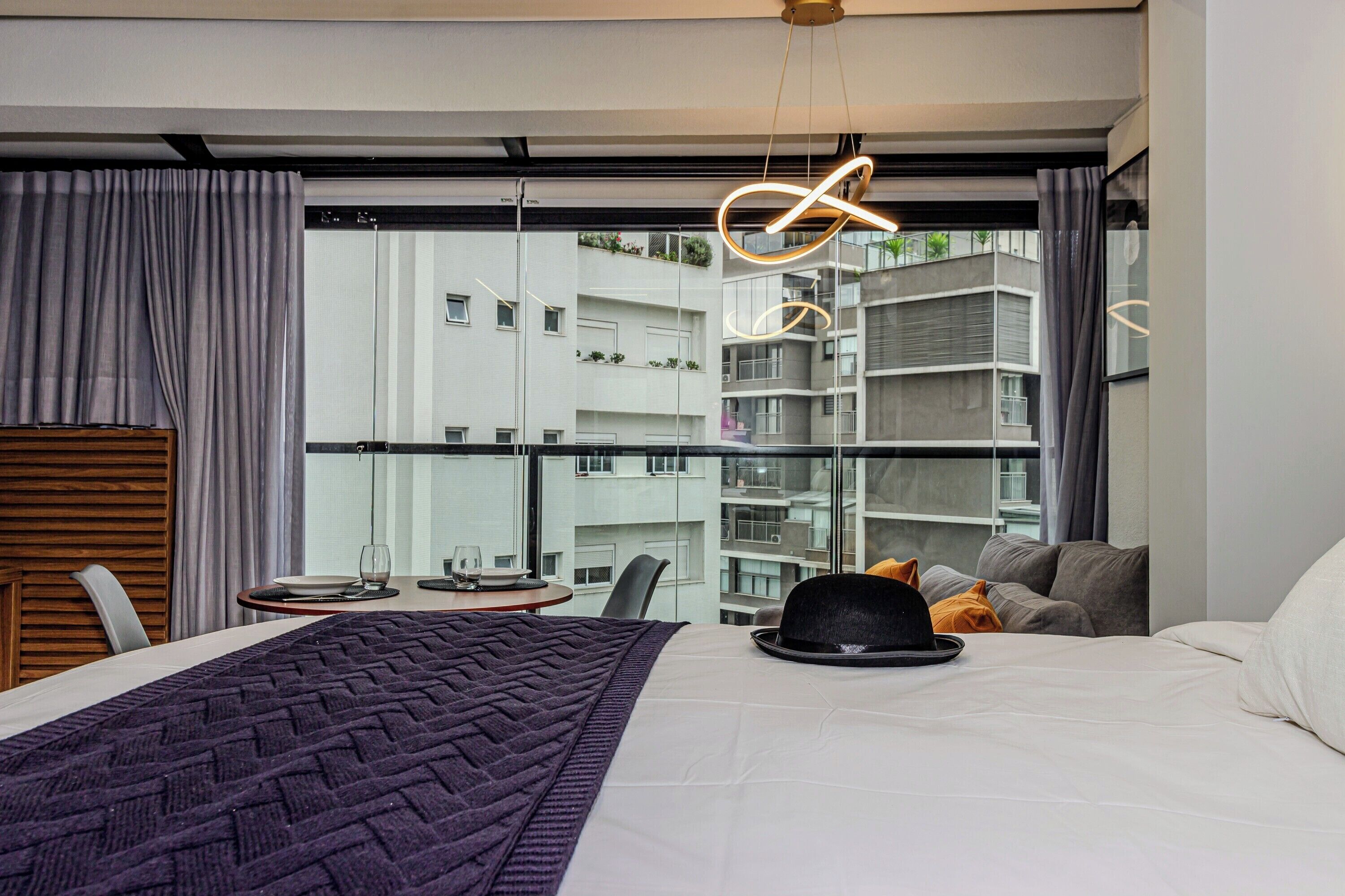 Quarto de hotel moderno com uma cama grande, uma mesa de jantar e vista para um edifício com varandas.