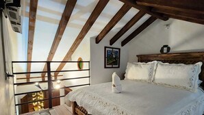 Quarto com teto de madeira, cama com lençóis brancos, uma mesinha e um quadro na parede.