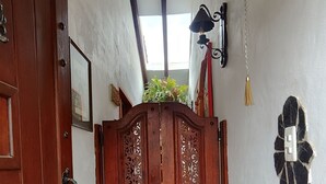 Um portão de madeira com detalhes entalhados e a letra 'H', dando acesso a um espaço de entrada com piso de azulejos e soalho de pedra.