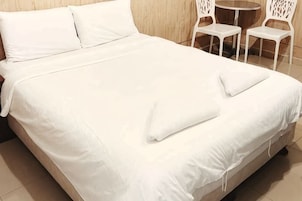 Cama bem arrumada com roupa de cama branca, uma mesinha redonda pequena e duas cadeiras brancas num quarto com paredes de madeira.